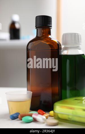 Bottles of syrup, measuring cup and pills on white table against light ...