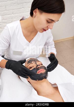 cosmetologist applying black mask on pretty woman face wearing black ...