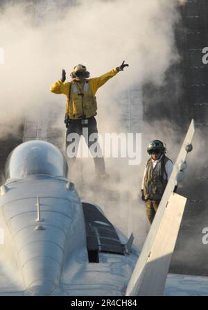 An aircraft handler directs an F/A-18 Hornet into position over a steam ...