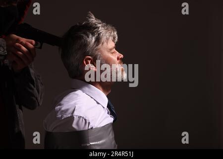 Kidnapper aiming gun at tied victim on dark background. Hostage taking ...