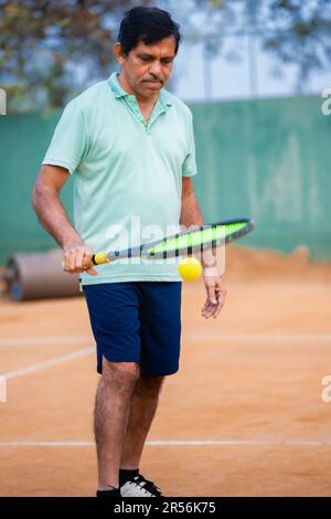 indian senior man practicing tennis by tapping on ground at court ...
