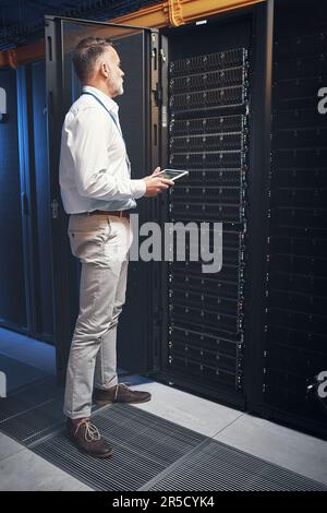Serious mature network engineer standing at open server cabinet and ...