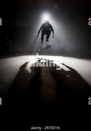 Young man kickflipping skateboard wearing all black, back lit with fog ...