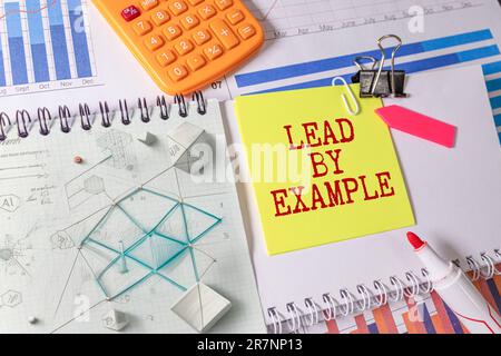 Text LEAD BY EXAMPLE on Office desk table with keyboard, notepad and analysis chart on a white ...