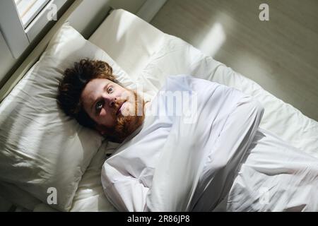 Young deranged man in straitjacket biting pillow while lying on bed by ...