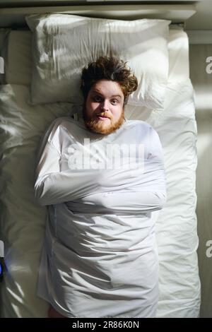 Young deranged man in straitjacket biting pillow while lying on bed by ...