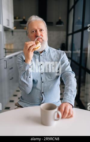 Disgruntled mature adult male eating unappetizing burger at kitchen ...