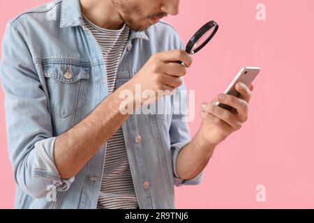 Handsome young man with magnifier and mobile phone on pink background ...