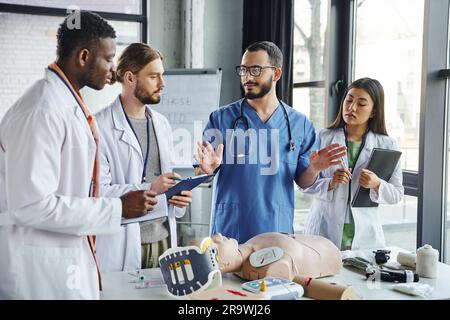 multiethnic students writing next to instructor showing wound care simulator near CPR manikin ...