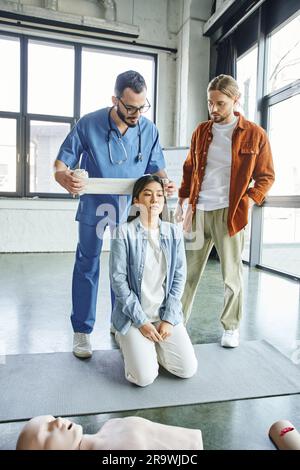 medical instructor in uniform and eyeglasses pointing at CPR manikin ...