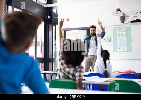 Diverse male teacher and pupils raising hands in elementary school ...