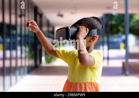 Biracial schoolgirl using vr headset reaching out hand in sunny school ...