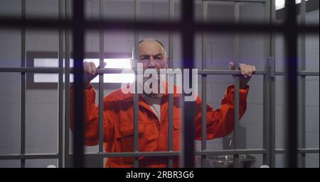 Elderly prisoner in orange uniform stretches his hands, leans on metal ...