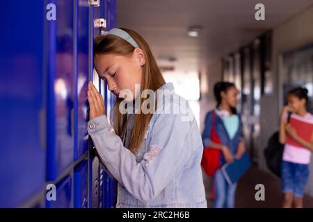 Sad caucasian schoolgirl standing next to lockers with diverse ...