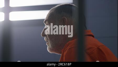 Elderly criminal in orange uniform sits in jail cell, stands up and ...