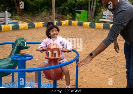 A father and daughter twirl on a spinner in a park. The father is ...