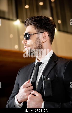 good looking bodyguard in sunglasses, handsome man in suit touching ...