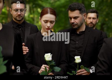 Funeral ceremony. Sad people with white rose flowers mourning outdoors ...