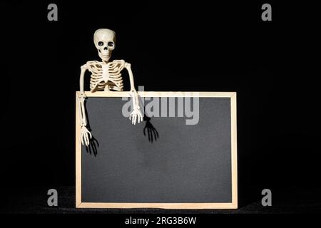 Funny human skeleton holding an empty black blank on a black background ...