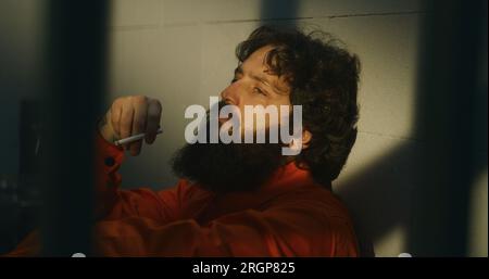 Thoughtful male prisoner in orange uniform sits on the bed, lights ...
