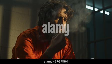 Depressed prisoner in orange uniform sits on bed in prison cell, smokes ...
