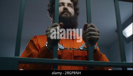 Prisoner in orange uniform holds metal bars, stands in prison cell in ...