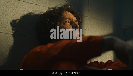 Depressed prisoner in orange uniform sits on bed in prison cell, smokes ...