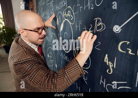 Young thoughtful man, scientist standing at blackboard with formulas ...