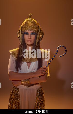 Brunette woman in egyptian attire and headdress holding crook on brown ...