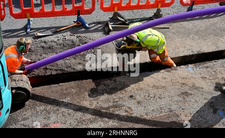 Workmen installing ducting for fibre optic cable in trench cut in road ...