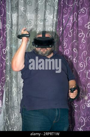 Overweight man playing virtual reality raising hands with joysticks at ...
