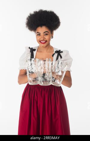 joyful african american bavarian waitress in dirndl holding beer mugs ...