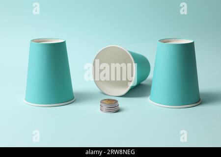 Three paper cups and coins on light blue background. Thimblerig game ...