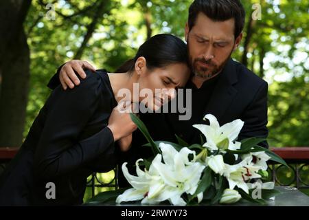 Sad couple mourning near granite tombstone with white lilies at ...