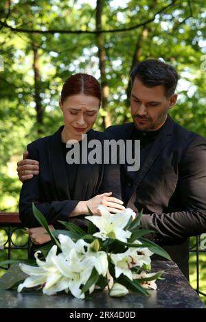 Sad couple mourning near granite tombstone with white lilies at ...