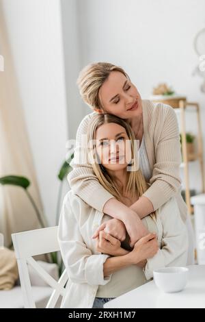 two trendy blonde sisters hugging warmly on white curtain background, family bonding, banner ...
