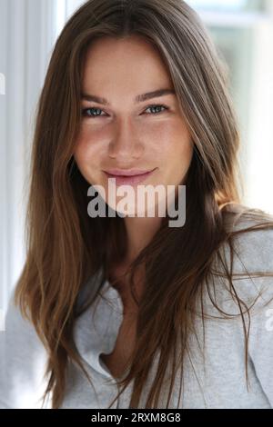 A smiling young woman with brown hair and a beige shirt leans against a ...