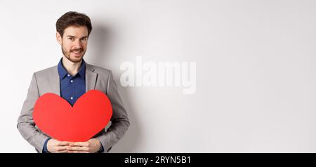 Handsome bearded man wishing happy valentines day, holding red heart ...