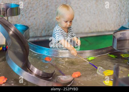 Toddler boy catching toy fish with a net in a vibrant water play area ...