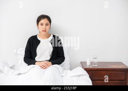 An uncomfortable woman sits on the bed and has medicine on the table ...