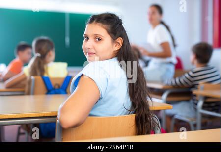 Positive tween schoolgirl sitting at school desk at lesson in class ...