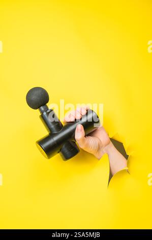 Female hand sticking out with massage gun from yellow paper background ...