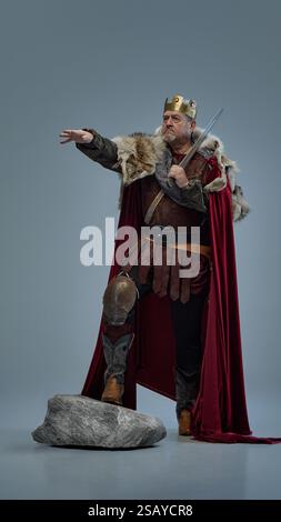 Senior serious man as medieval king standing on stone, leaning on sword ...