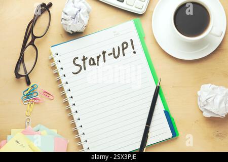 Start up word on notebook page with calculator and coffee on blue table ...