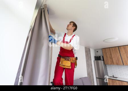 A male repairman installs a curtain rod and hangs curtains in the house ...