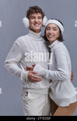 multiethnic couple in knitted sweaters and with snowy hair looking at ...