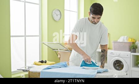 Young hispanic man ironing clothes holding folded clothes at laundry ...