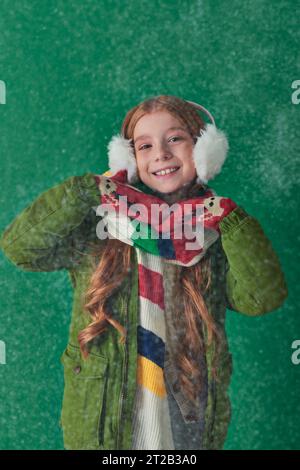 positive child in ear muffs and scarf standing in winter outfit on ...