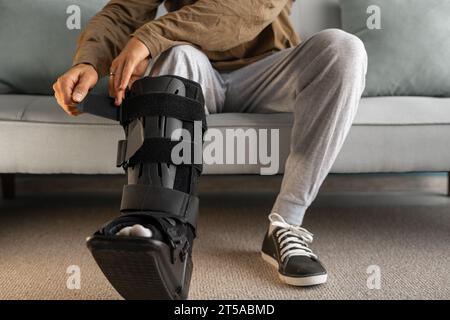 Man putting ankle sprain orthosis, black splint on leg sitting on the sofa on white background ...