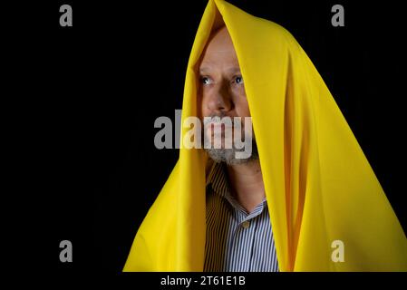 Bald, bearded man with a yellow cloth over his head against black ...